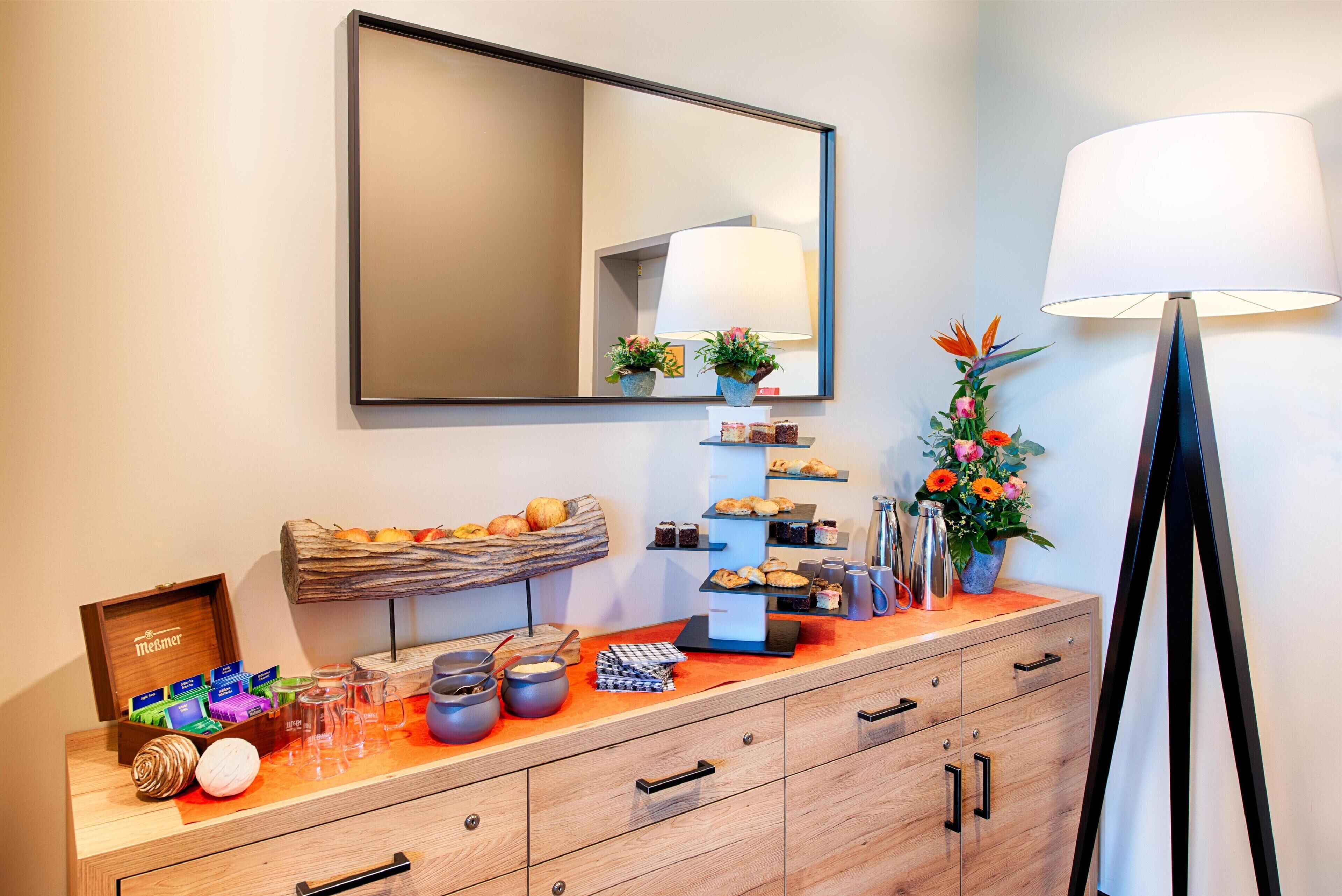 Quarto de hotel com cômoda de madeira, um espelho, uma luminária e uma mesinha com diversos itens, incluindo uma bandeja de doces, um vaso com flores e uma caixa de chocolates.