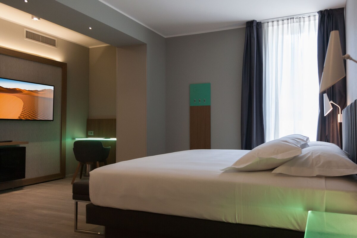Quarto de hotel com cama, escrivaninha, televisão e janela com cortinas.