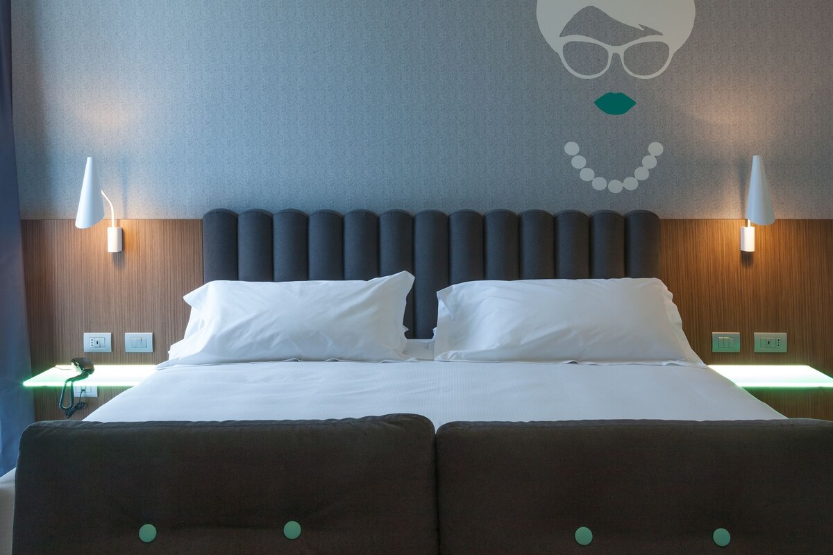 Uma cama de casal com roupa de cama branca, uma cabeceira escura e duas luminárias de apoio.