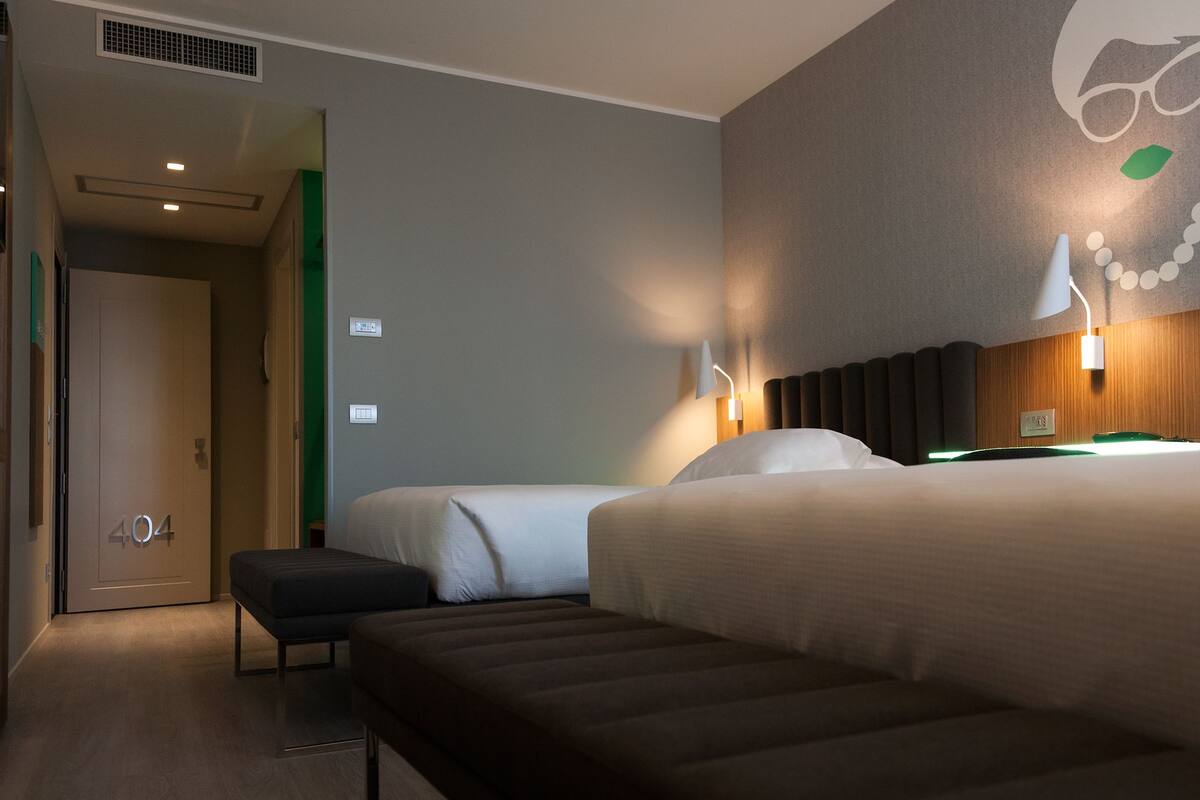 Quarto de hotel com duas camas, um banquinho, uma luminária na parede e uma porta com número.