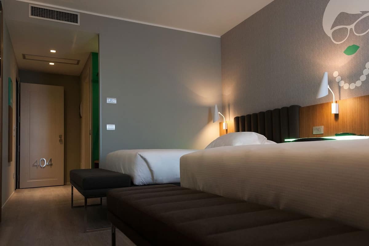 Quarto de hotel com duas camas, um banco, uma luminária na parede e uma porta com número.