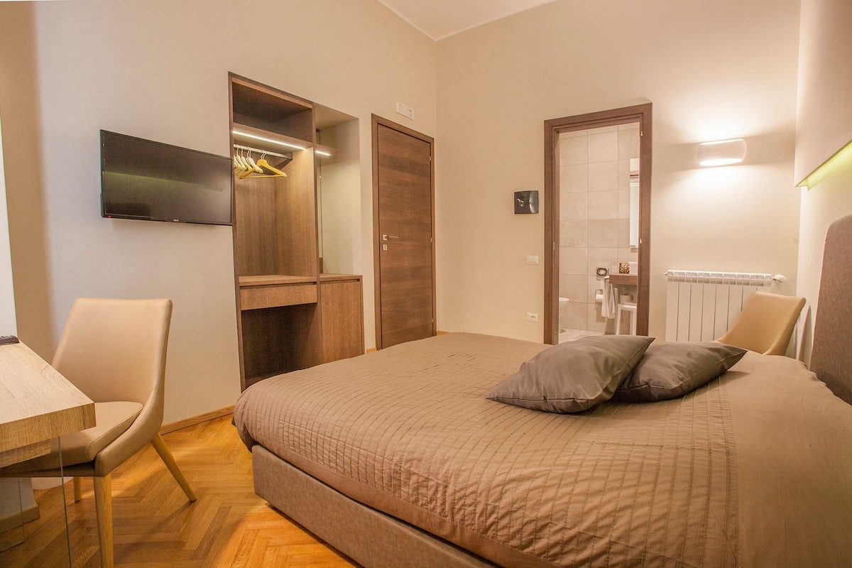 Um quarto com cama, escrivaninha, cadeira, guarda-roupa e televisão.