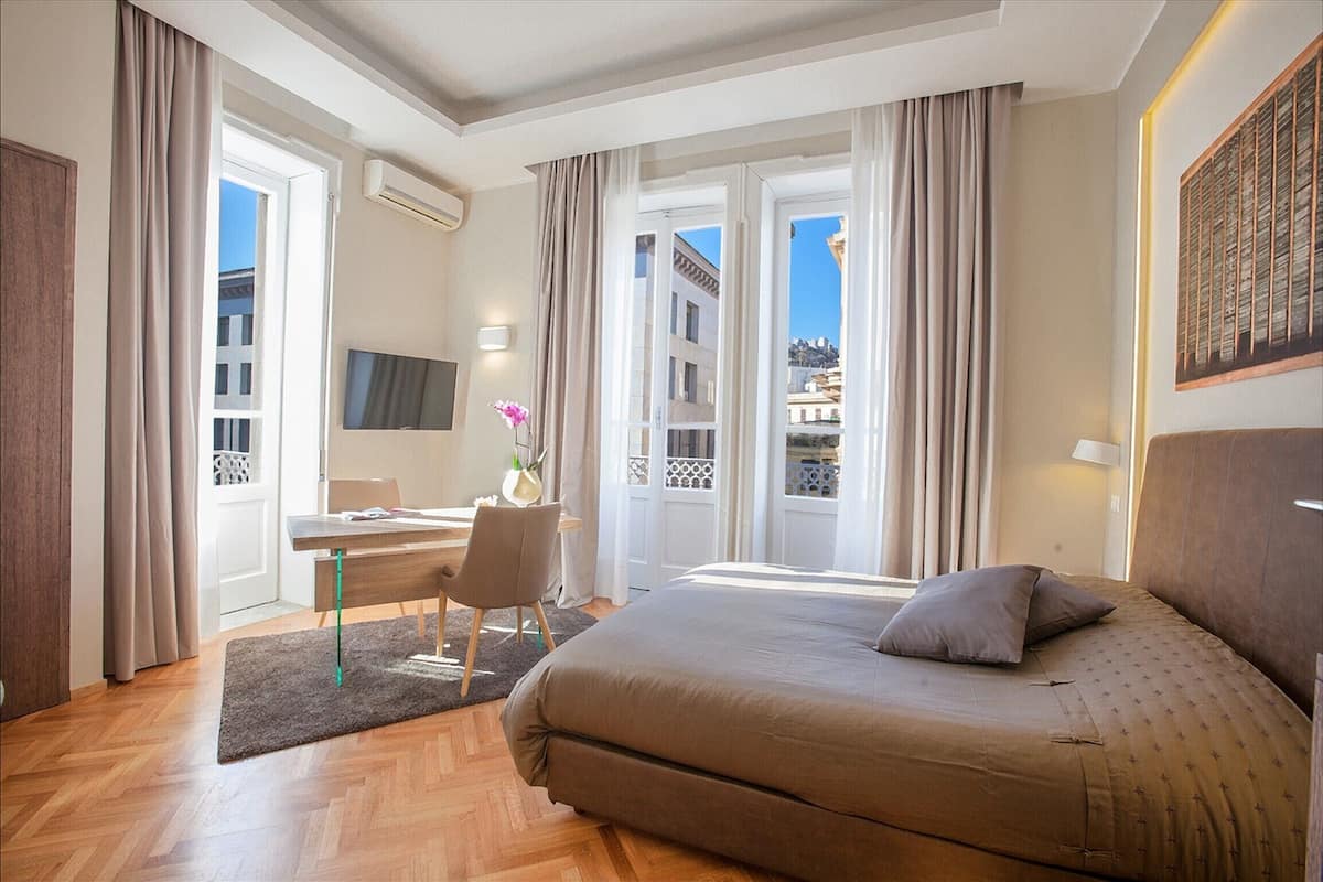 Quarto de hotel com cama, escrivaninha, cadeira e uma janela grande com cortinas.