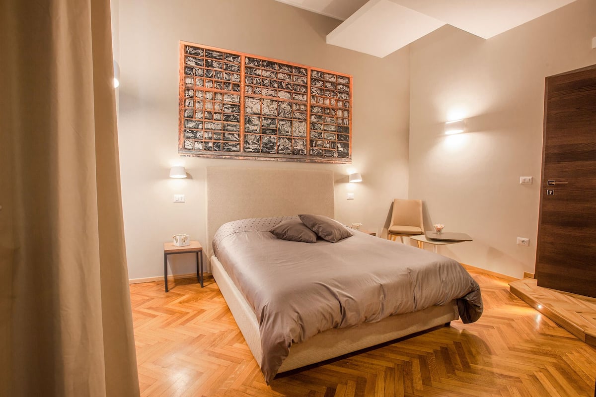 Quarto com cama, uma mesinha, cadeira e piso de madeira.