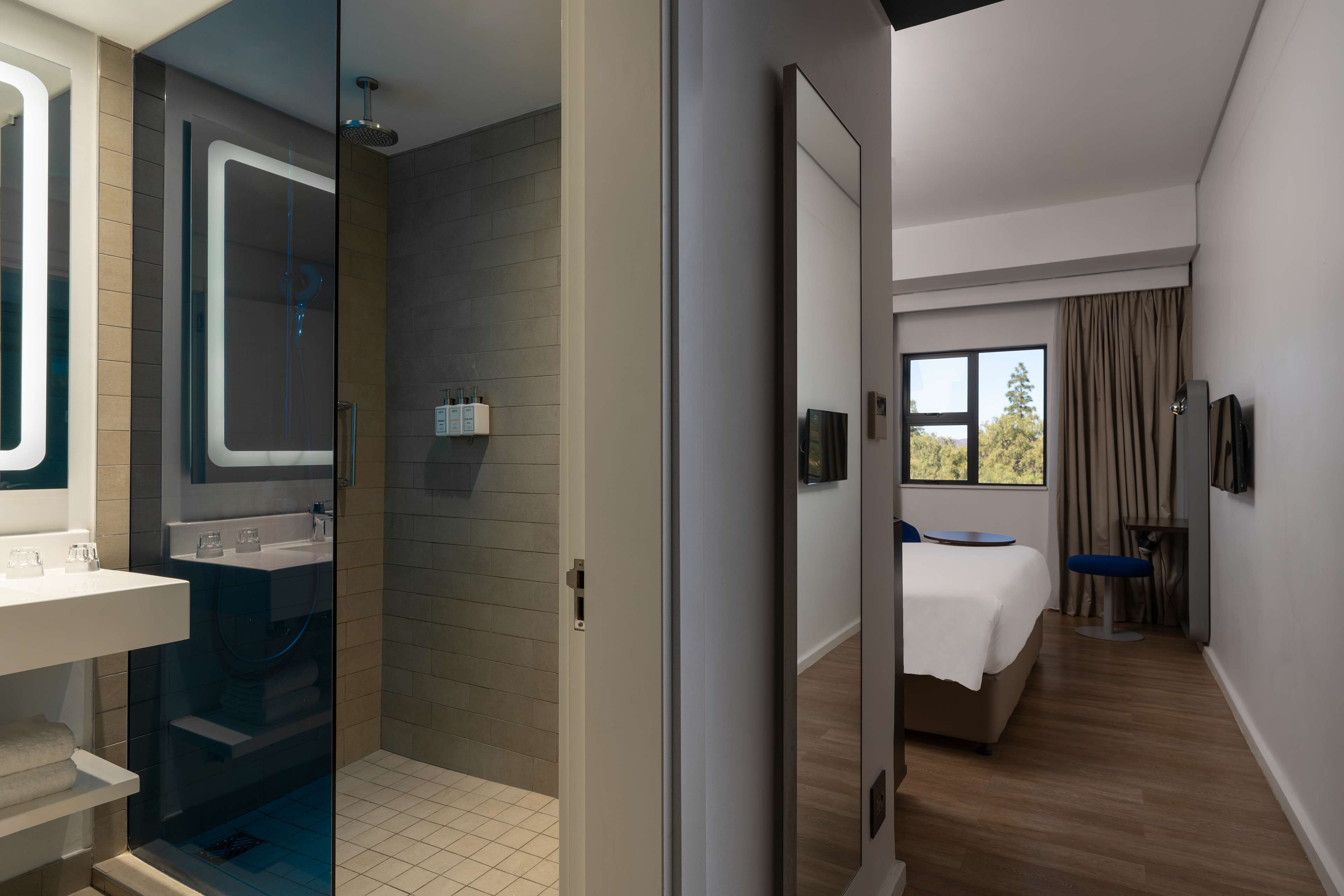 Quarto de hotel moderno com chuveiro em cabine de vidro, uma cama grande, mesa de cabeceira e vista para o exterior.