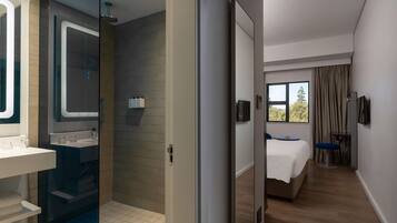 Quarto de hotel moderno com chuveiro em cabine de vidro, uma cama grande, mesa de cabeceira e vista para o exterior.