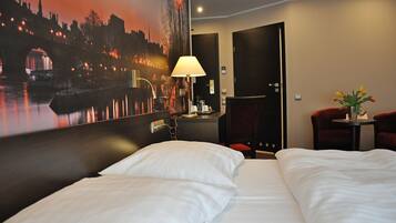 Quarto de hotel com cama, secretária com candeeiro, cadeira, pequena mesa com um vaso e um quadro de uma paisagem urbana.