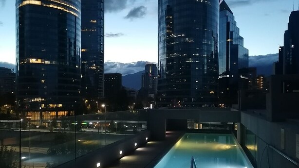 Piscina no terraço com barreira de vidro, rodeada por edifícios modernos e altos.