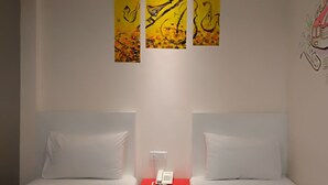 Quarto de hotel com duas camas, mesa de cabeceira vermelha com telefone e três quadros amarelos na parede.
