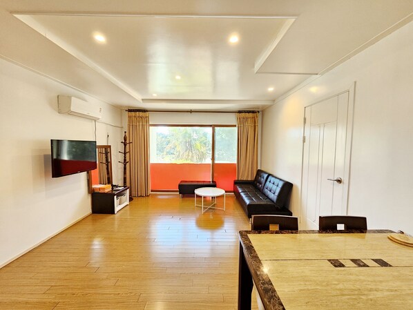 Uma sala de estar ampla com piso de madeira, um sofá preto, uma mesinha redonda e uma TV fixada na parede.