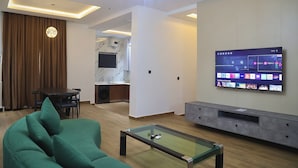 Sala de estar moderna com um sofá verde, uma mesa de centro de vidro, uma TV de tela plana na parede e uma área de cozinha ao fundo.