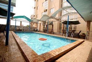 Uma piscina com um salva-vidas, rodeada por espreguiçadeiras e guarda-sóis, e um edifício ao fundo.