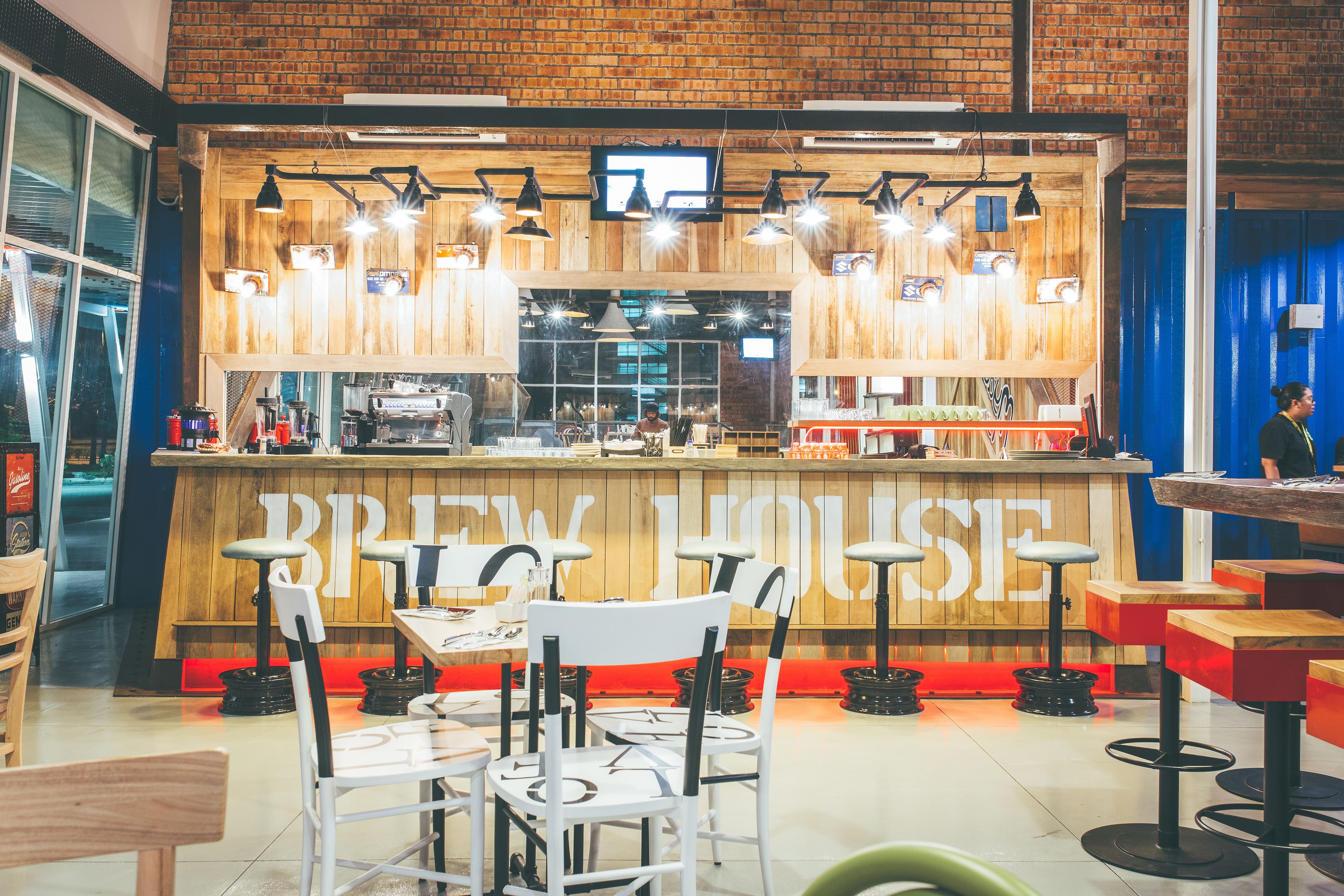 Interior de uma cafeteria com balcão de madeira identificado como 'BREW HOUSE', várias mesas e cadeiras, e uma parede de tijolos ao fundo.