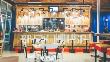 Interior de uma cafeteria com balcão de madeira identificado como 'BREW HOUSE', várias mesas e cadeiras, e uma parede de tijolos ao fundo.
