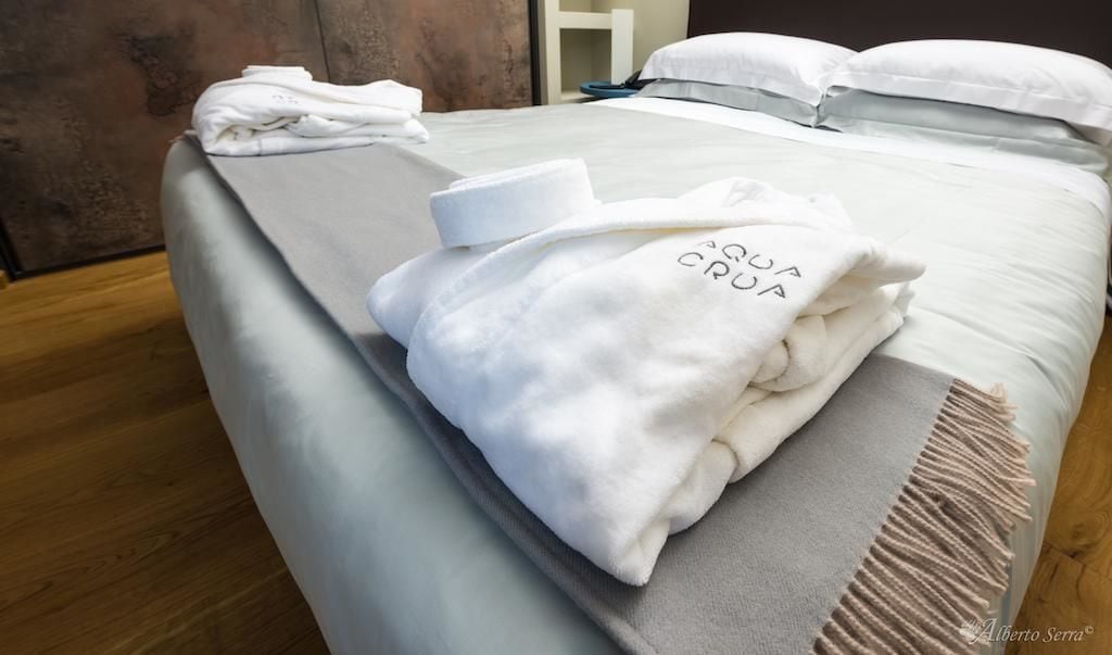 Cama bem arrumada com lençóis brancos e uma toalha dobrada com texto, piso de madeira e parede lisa.
