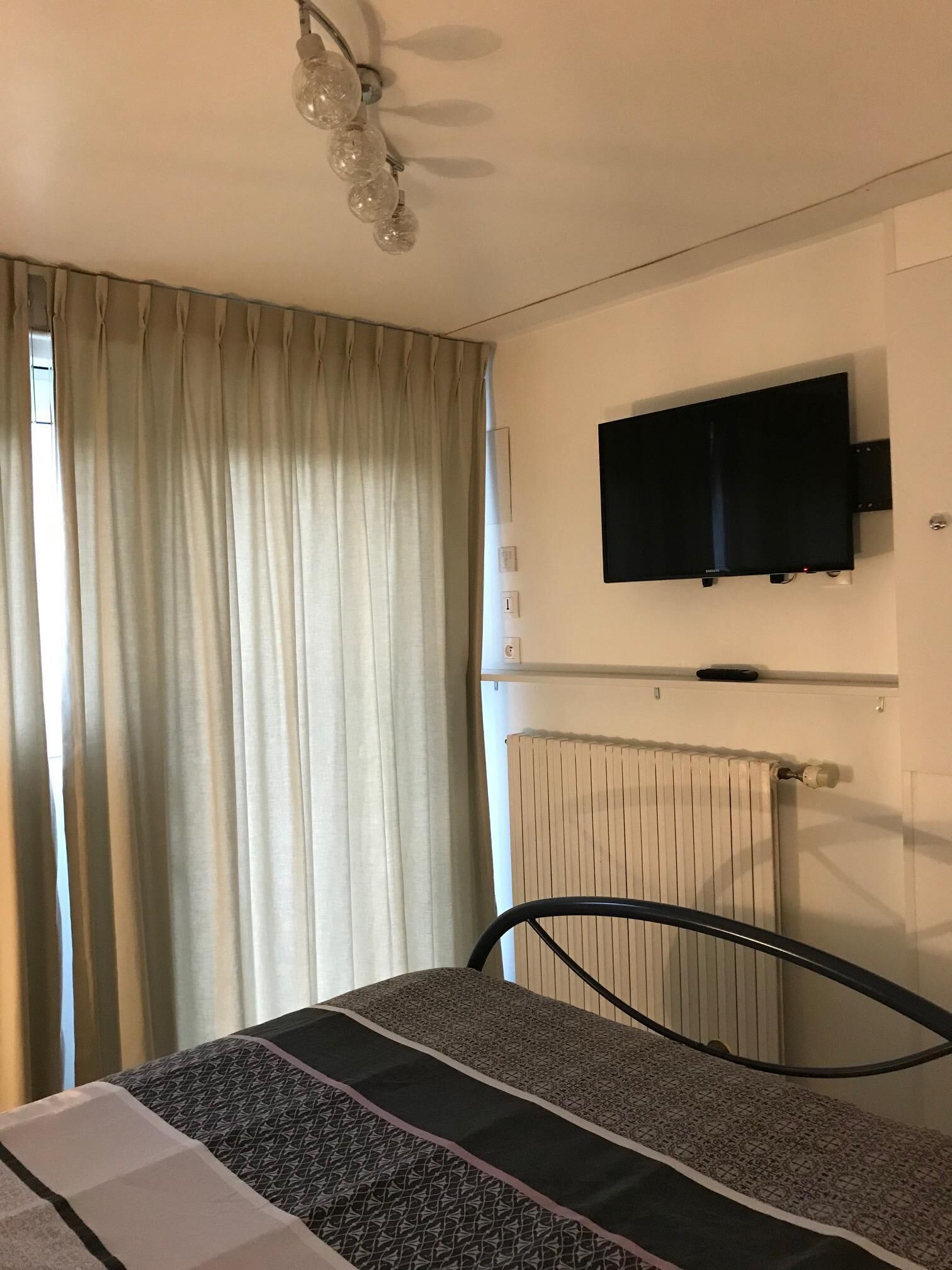 Quarto com cama, televisão na parede, radiador e janela com cortinas.