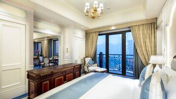 Quarto de hotel espaçoso com uma cama grande, área de estar com uma poltrona e uma mesa, varanda com vista e um lustre.