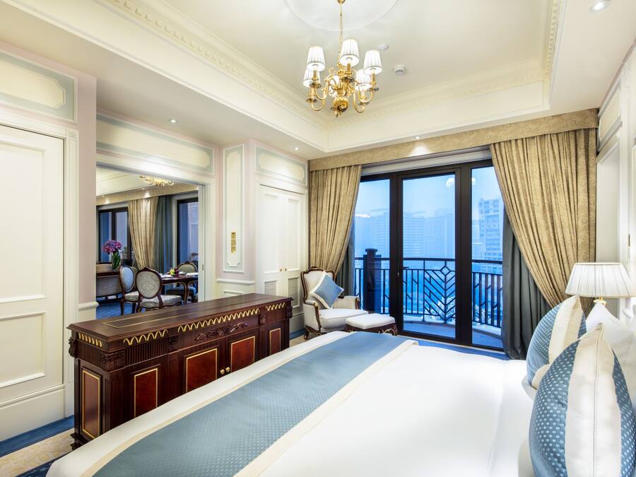 Quarto de hotel espaçoso com uma cama grande, área de estar com uma poltrona e uma mesa, varanda com vista e um lustre.