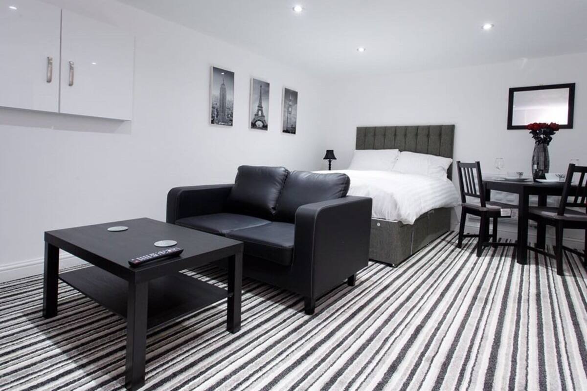 Quarto de hotel moderno com uma cama, um sofá de couro preto, uma mesinha e um espaço de jantar com cadeiras.