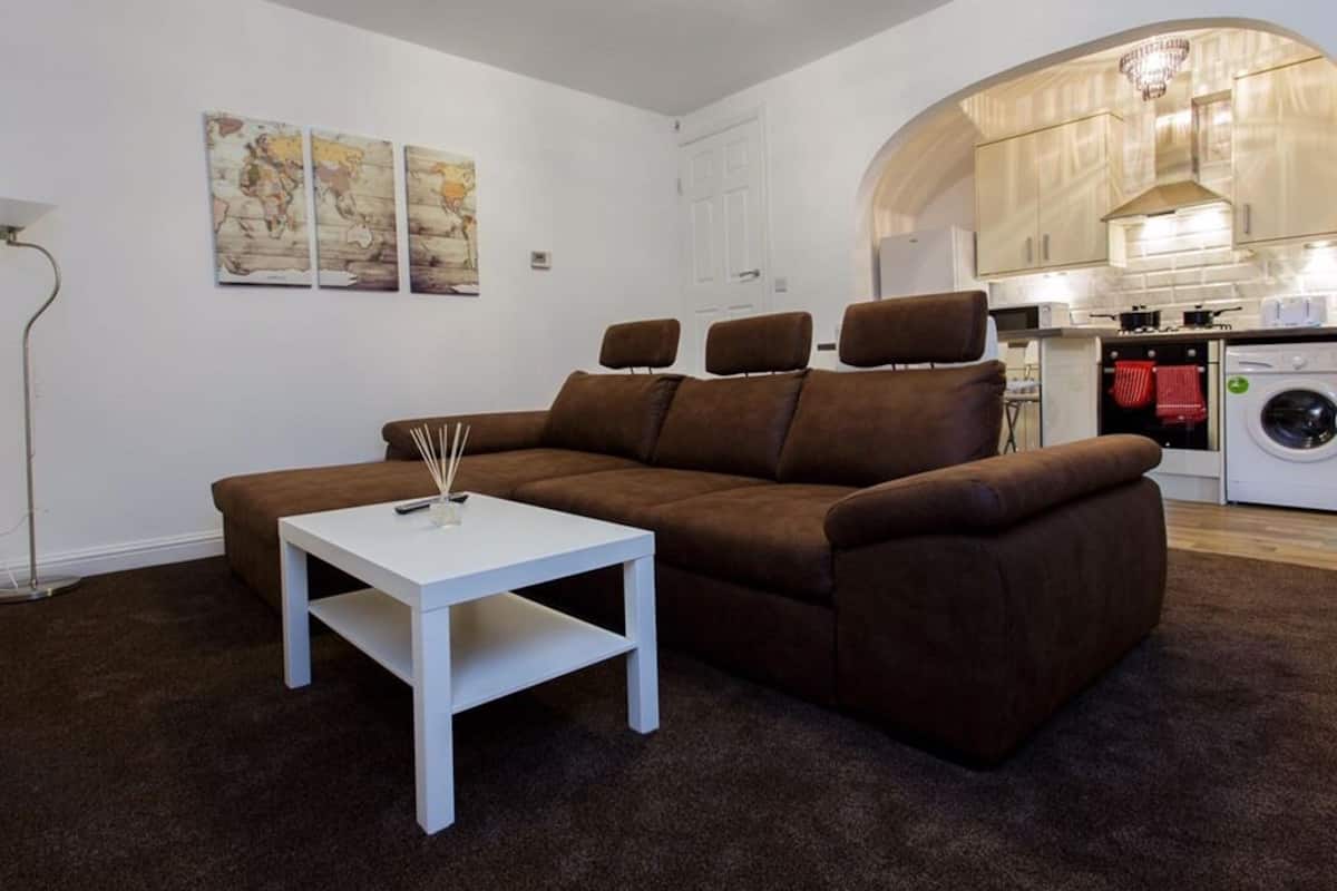 Uma sala de estar moderna com um sofá seccional marrom, uma mesa de centro branca e uma pequena mesa lateral branca. Cozinha visível ao fundo, com eletrodomésticos de aço inoxidável e um lustre.