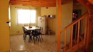 Uma sala de jantar com mesa e cadeiras, geladeira, micro-ondas e uma janela com cortinas.