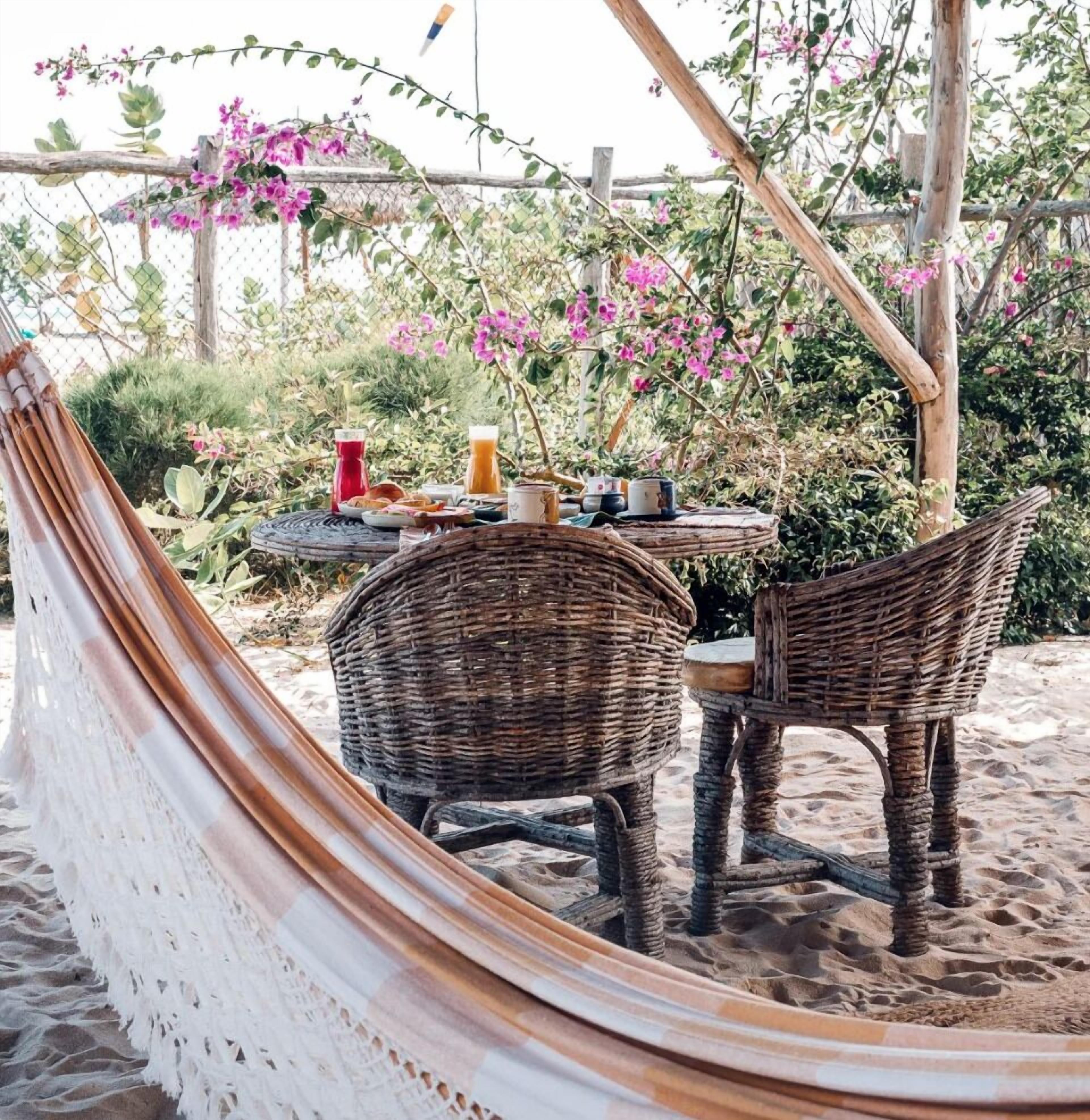 Uma rede montada em uma praia arenosa, com uma mesa e cadeiras ao fundo.