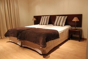 Uma cama bem arrumada com cabeceira marrom, duas mesas de cabeceira com luminárias e piso de madeira.