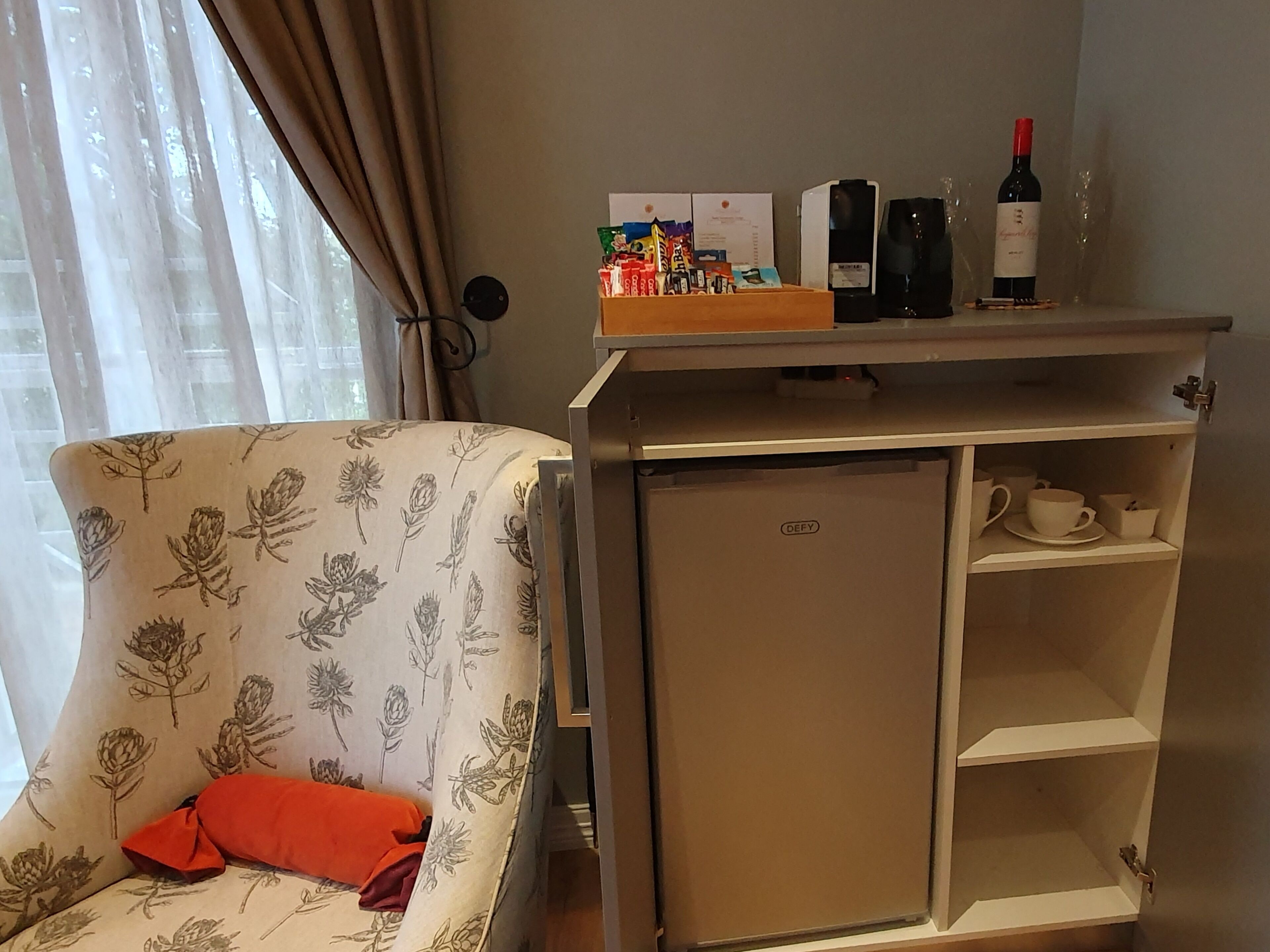Quarto com poltrona de padrão floral, um pequeno armário de madeira com uma garrafa de vinho e copos, e uma janela com cortinas.