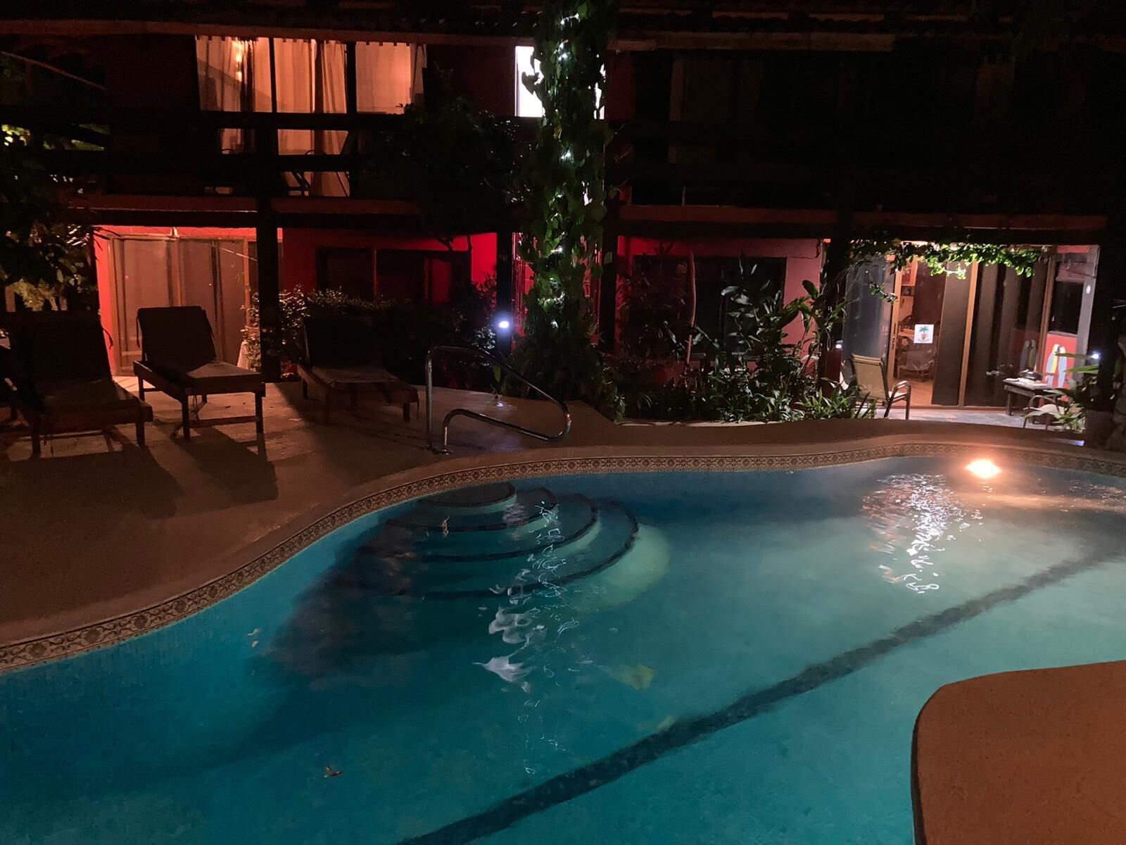 Uma piscina com espreguiçadeiras e hidromassagem, cercada por plantas e um edifício com janelas.