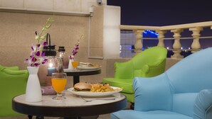 Uma área de refeições com uma mesa posta para dois, exibindo um hambúrguer, batatas fritas e um copo de suco de laranja. Há duas cadeiras, uma verde e outra azul, e um vaso com flores roxas.