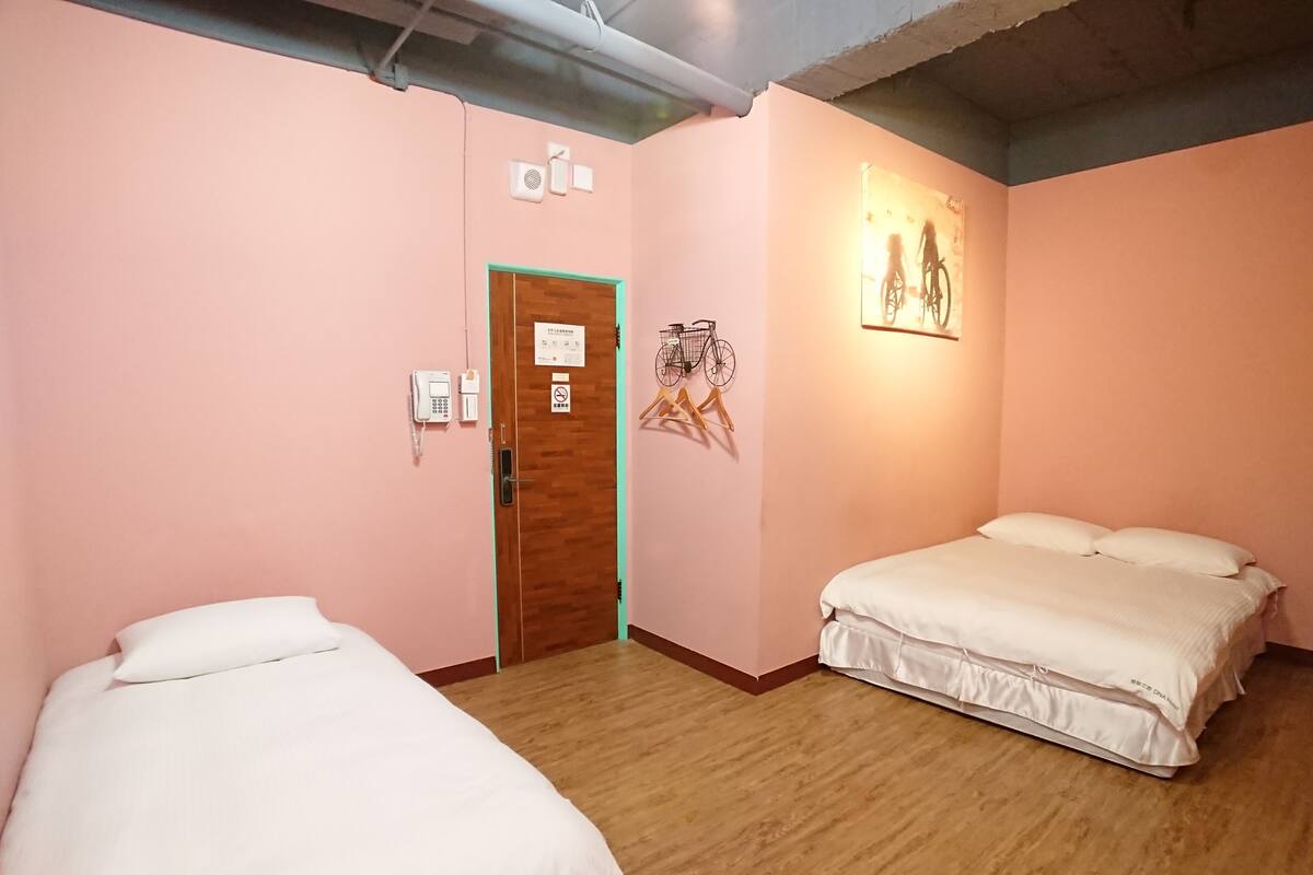 Quarto de hotel com duas camas, piso de madeira, porta com uma placa, telefone fixado na parede e uma obra de arte com tema de bicicleta.