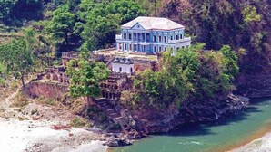 Um edifício grande, de vários andares, com fachada azul e telhado vermelho, rodeado por vegetação exuberante e um rio.