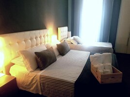 Quarto de hotel com uma cama grande, duas mesas de cabeceira e uma caixa de madeira com produtos de higiene pessoal.