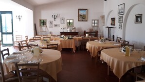 Uma sala de jantar com mesas redondas cobertas por toalhas bege, cadeiras de madeira e centros de mesa com flores.