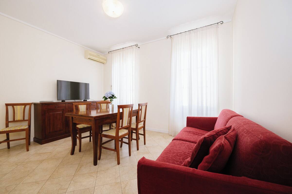 Uma sala de estar com sofá vermelho, mesa de jantar de madeira, cadeiras, televisão e janela com cortinas.