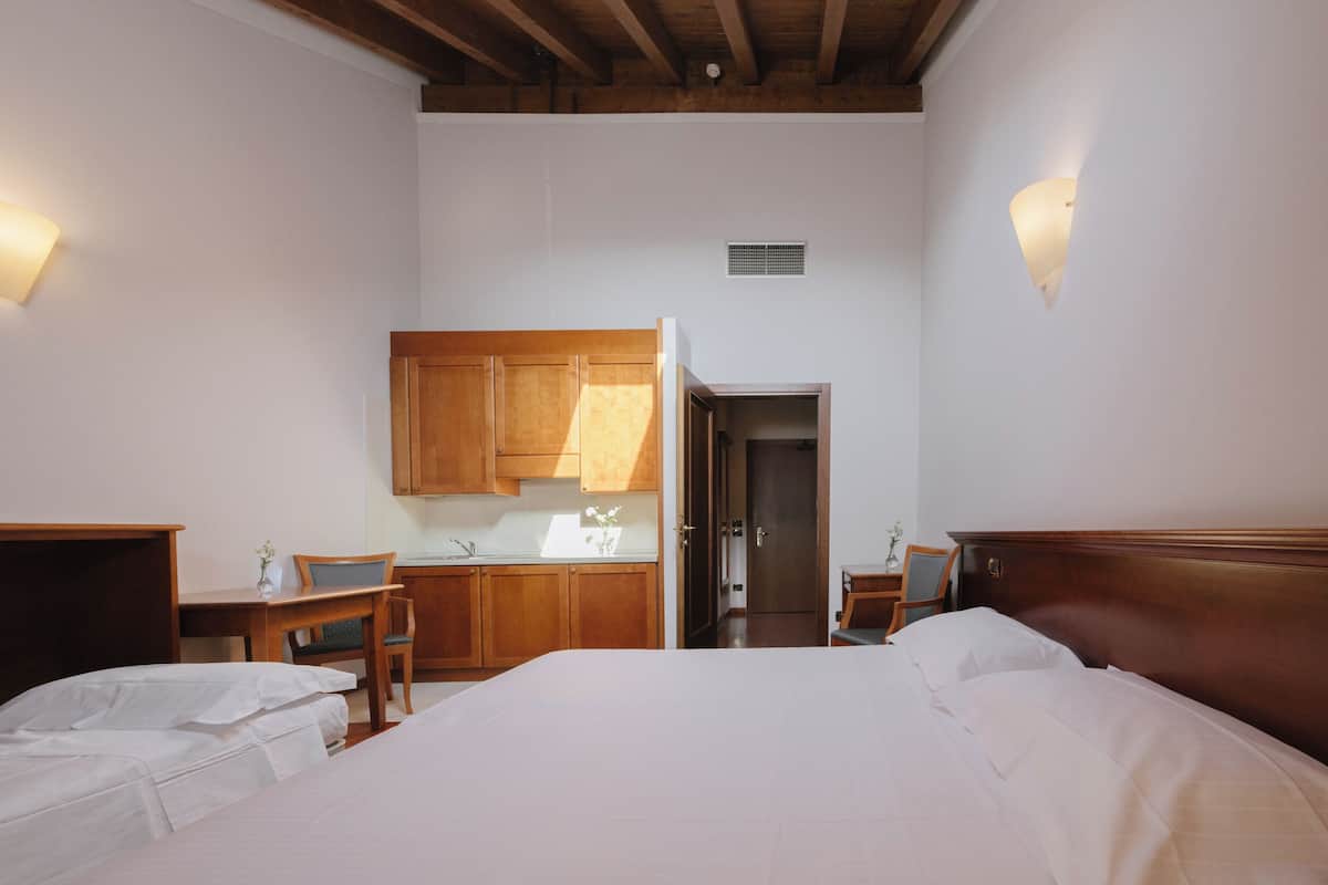 Quarto com cama de madeira, uma pequena escrivaninha e um espaço com cozinha compacta.