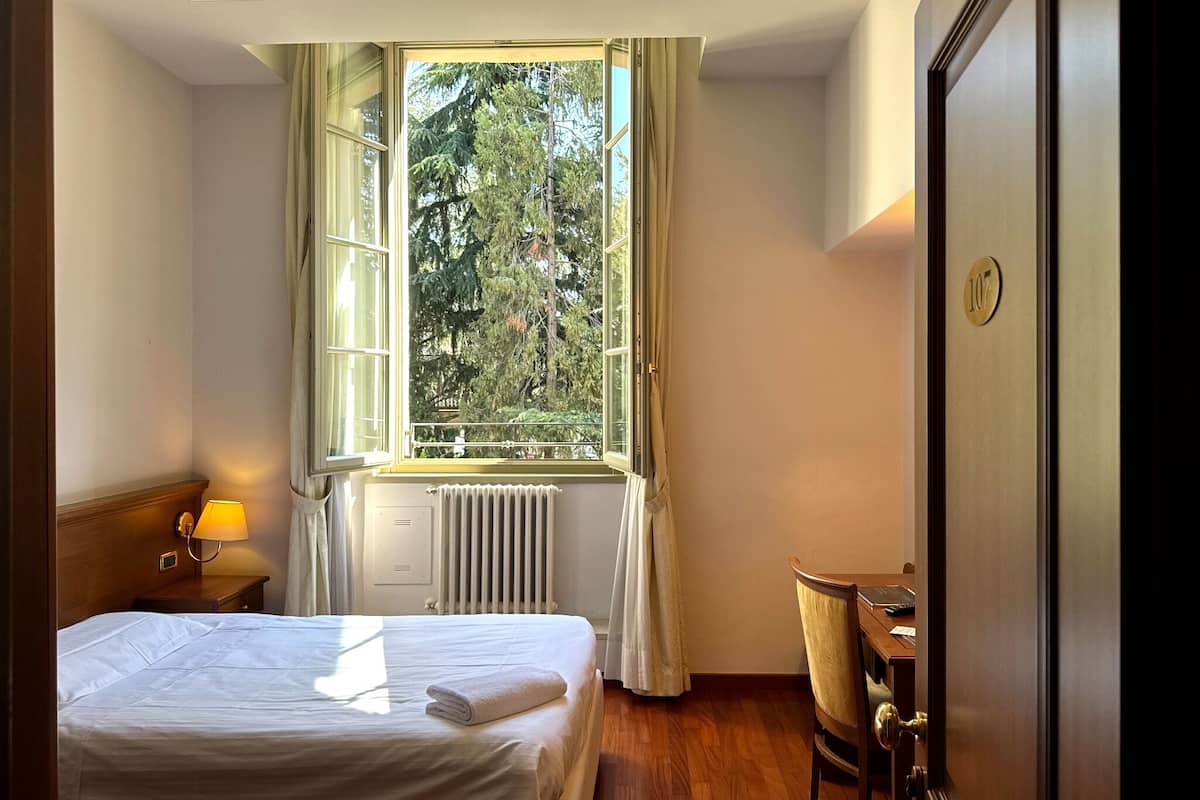 Quarto de hotel com cama, escrivaninha, janela com cortinas, radiador e porta com placa de identificação.