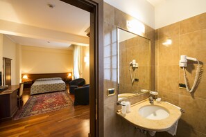 Quarto de hotel com cama, escrivaninha, cadeira, banheiro com pia e espelho, e chuveiro.