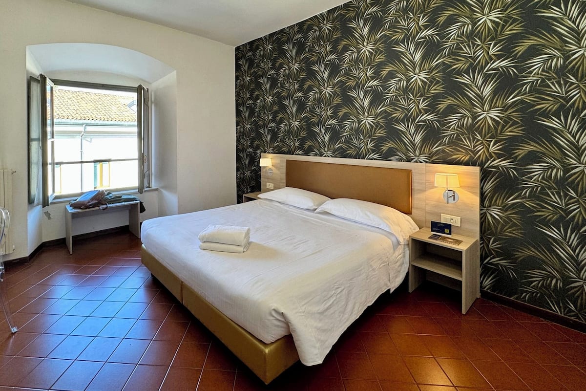 Quarto de hotel com cama, mesa de cabeceira, luminária, um banco e uma janela.
