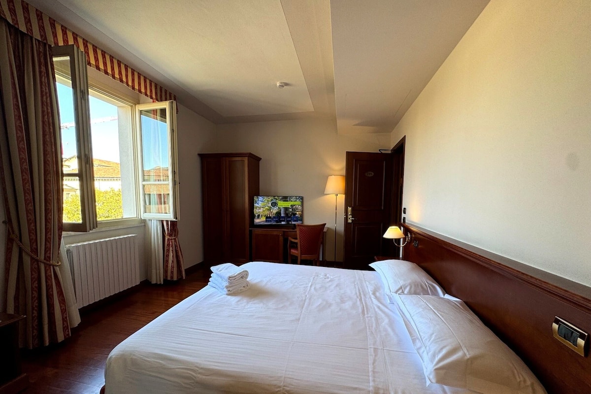 Quarto de hotel com uma cama, uma escrivaninha com cadeira, televisão e guarda-roupa.