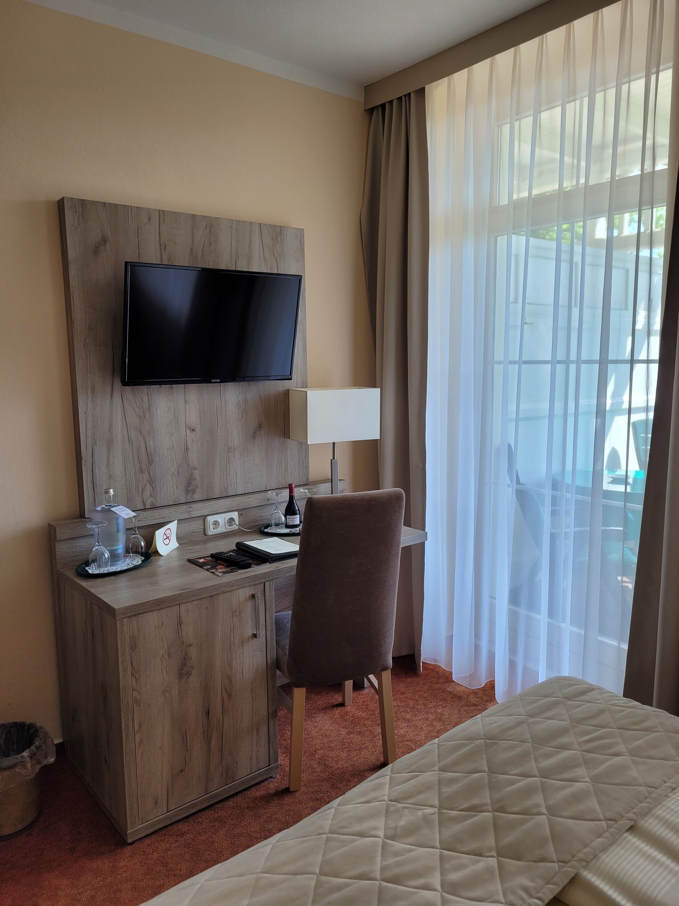 Quarto de hotel com escrivaninha de madeira, televisão de tela plana, cadeira, cama e janela com cortinas.