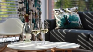Uma mesa com uma garrafa e duas taças de vinho branco, uma cadeira branca e um sofá com almofada florida.