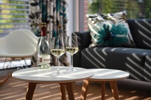 Uma mesa com uma garrafa e duas taças de vinho branco, uma cadeira branca e um sofá com almofada florida.