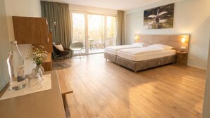 Quarto de hotel com cama, escrivaninha com garrafa e copos, uma cadeira, varanda com vista e um quadro na parede.