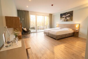 Quarto de hotel com cama, escrivaninha com garrafa e copos, uma cadeira, varanda com vista e um quadro na parede.