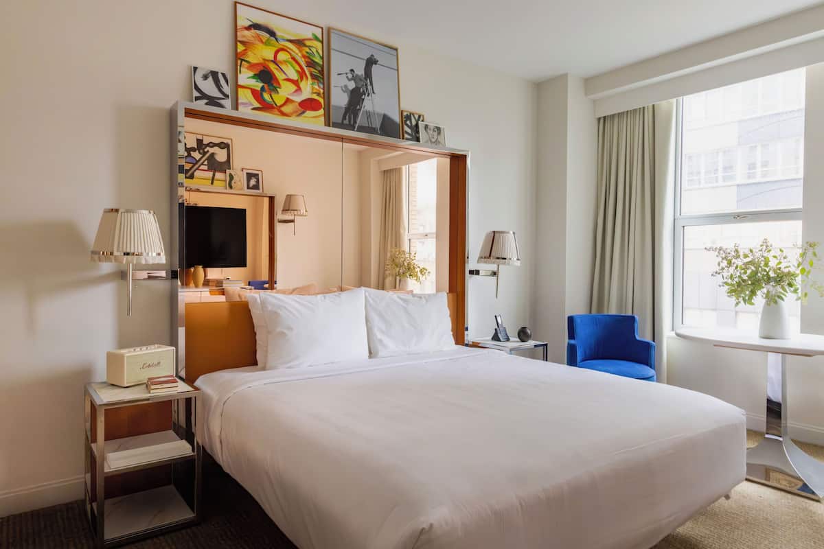 Quarto de hotel com uma cama grande, uma escrivaninha com luminária, uma cadeira, televisão e obras de arte na parede.