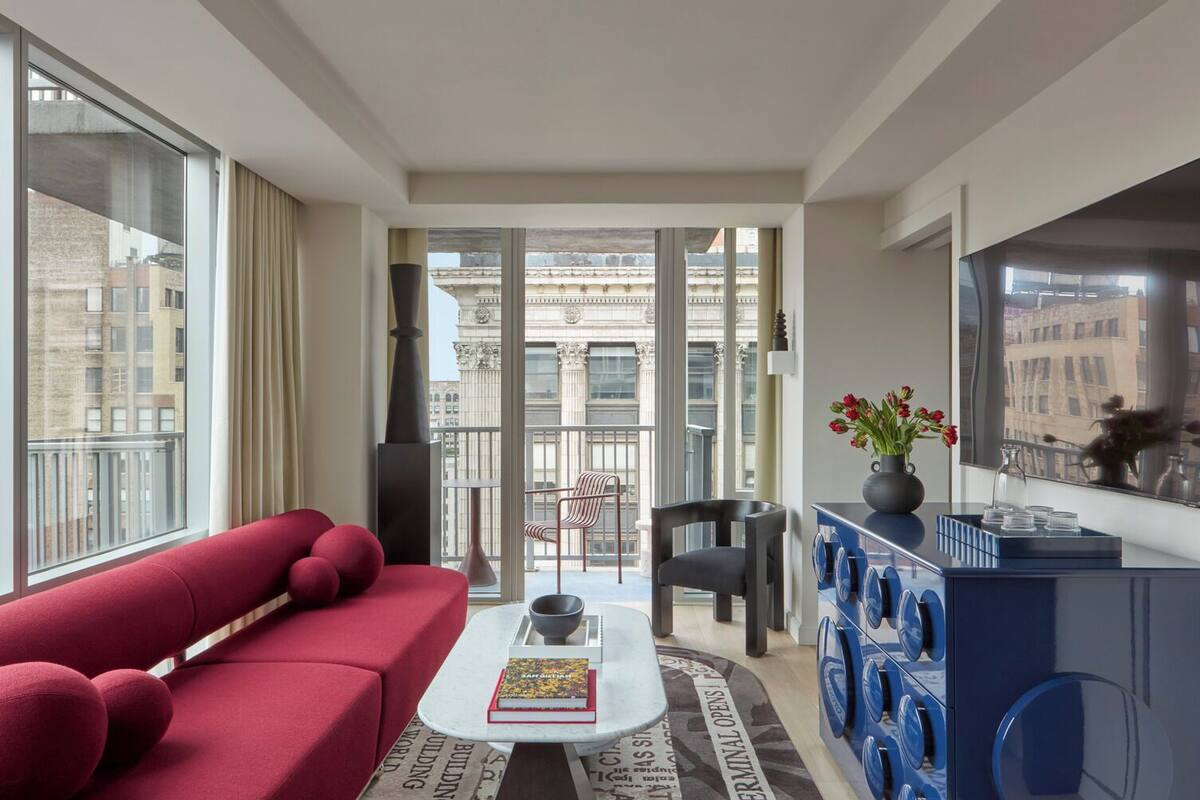 Uma sala de estar moderna com um sofá vermelho, uma mesa de centro com um livro, um armário azul e vista para um edifício através de grandes janelas.