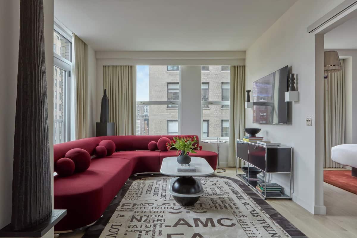 Quarto de hotel moderno com sofá seccional vermelho, mesa de centro redonda com um vaso, TV montada na parede e vista para a paisagem urbana através de grandes janelas.