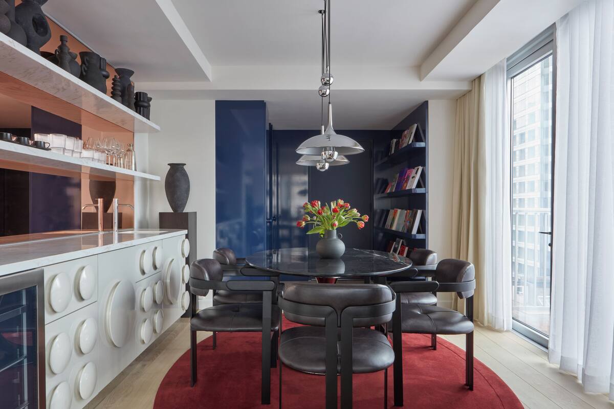 Uma área de refeições moderna com uma mesa redonda, cadeiras pretas, um vaso central com flores e uma luminária pendente.