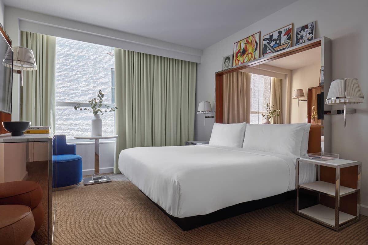 Quarto de hotel com uma cama grande, uma escrivaninha com luminária, uma cadeira, uma mesinha com um vaso e obras de arte na parede.